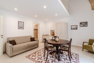 Uma sala de estar com sofá, uma mesa de jantar com cadeiras, um tapete estampado e uma área de cozinha com armários.