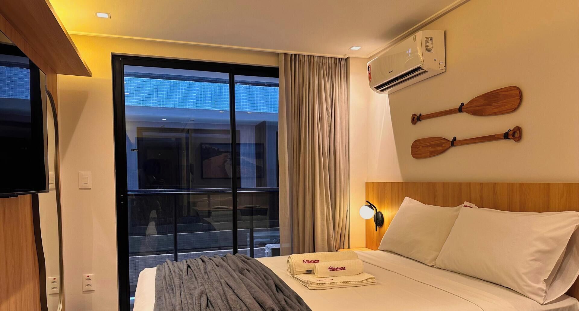 Quarto de hotel com cama, televisão, ar condicionado, janela com cortinas e vista para o exterior.