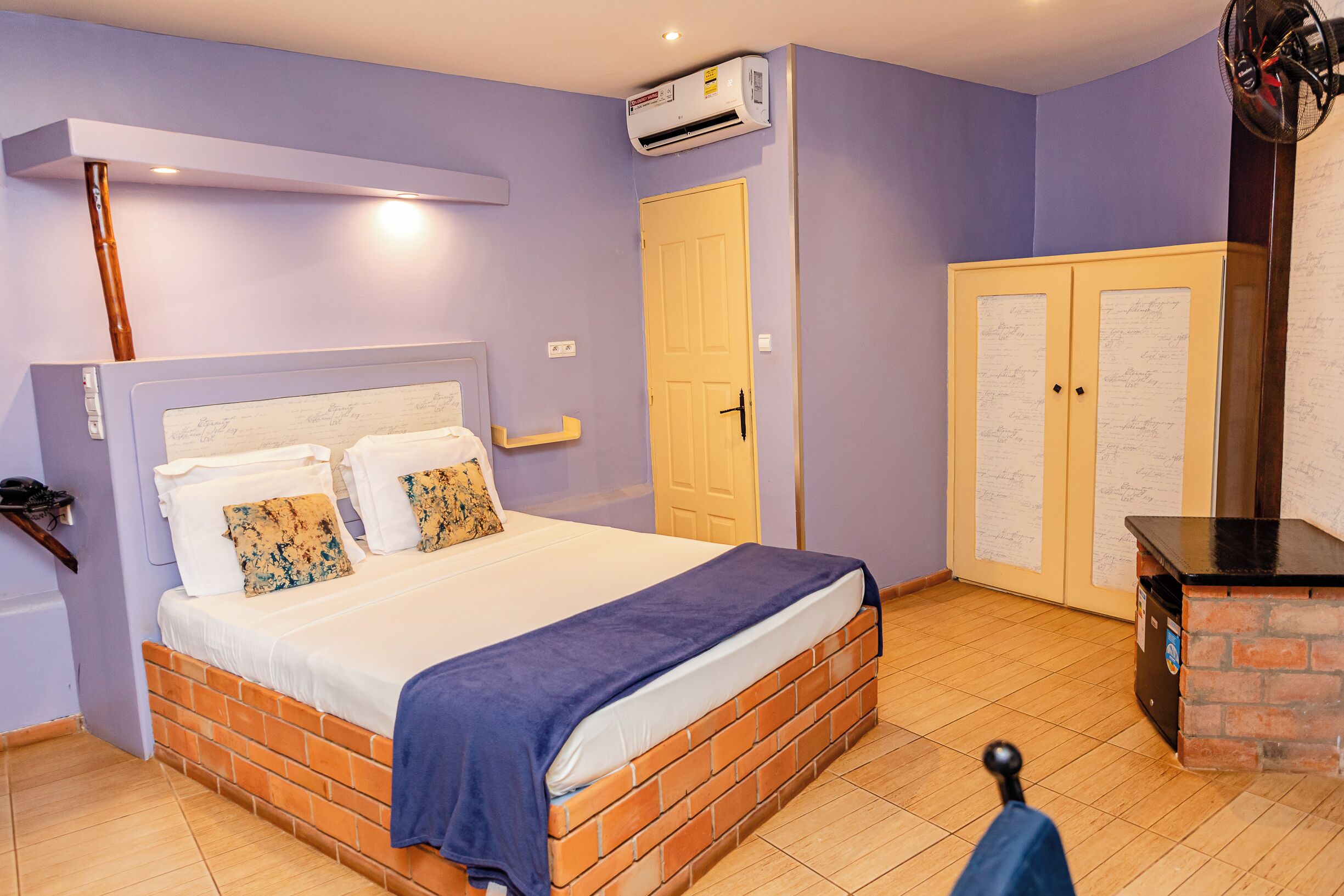 Quarto com cama tendo cabeceira de tijolos, manta azul, guarda-roupa de madeira e pequena lareira de tijolos.