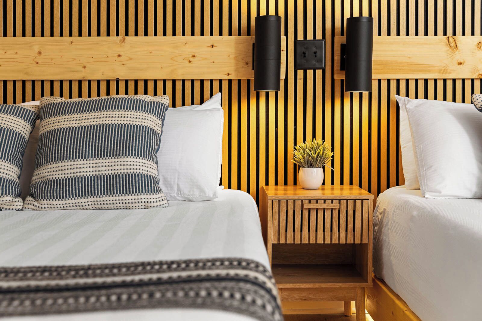 Quarto de hotel com parede de ripas de madeira, cama com roupa de cama listrada, mesa de cabeceira com uma planta e mesa de apoio com abajur.
