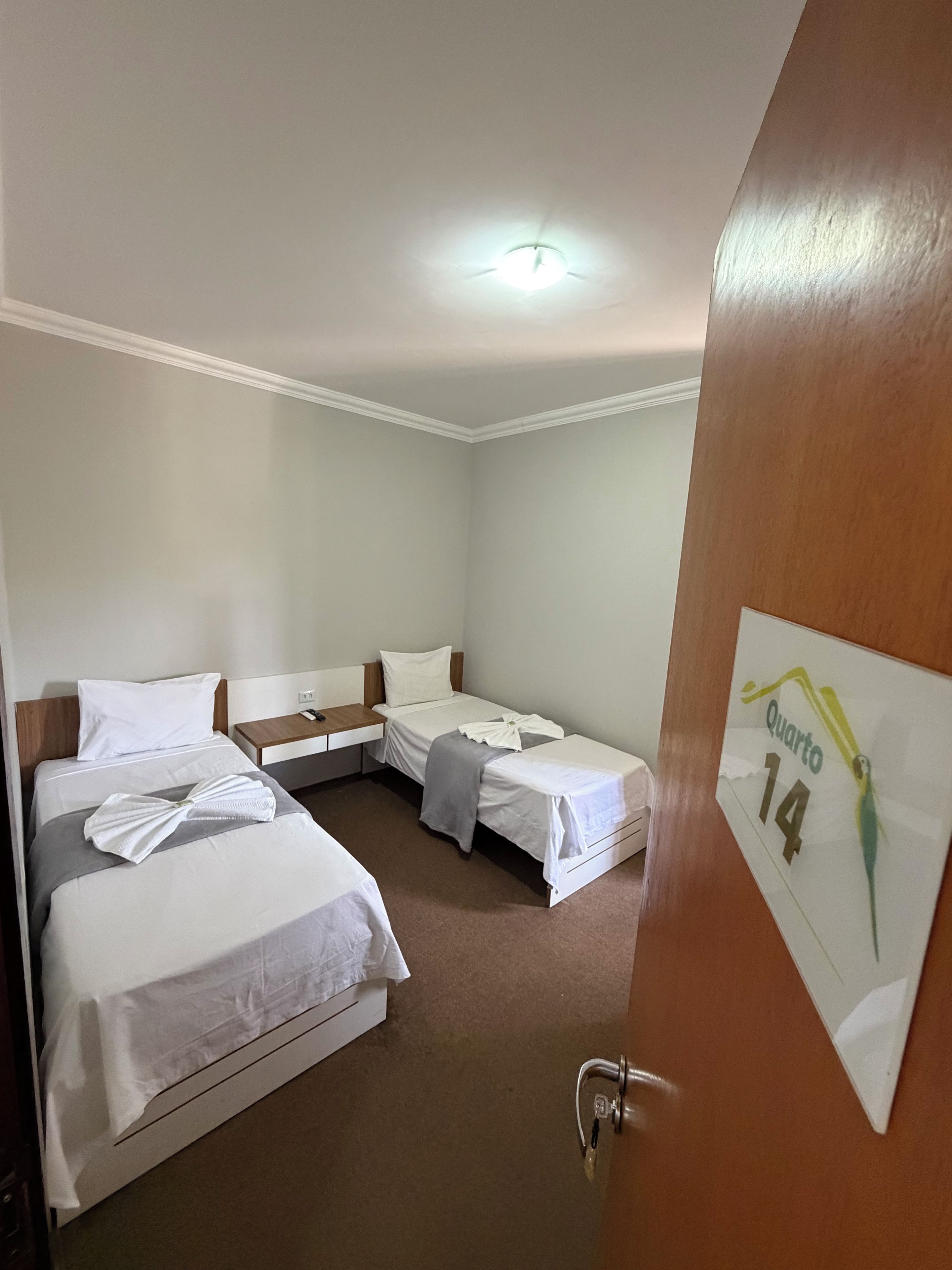 Quarto de hotel com duas camas de solteiro, uma mesa de cabeceira de madeira, luminária fixada na parede e uma porta com o símbolo de número.