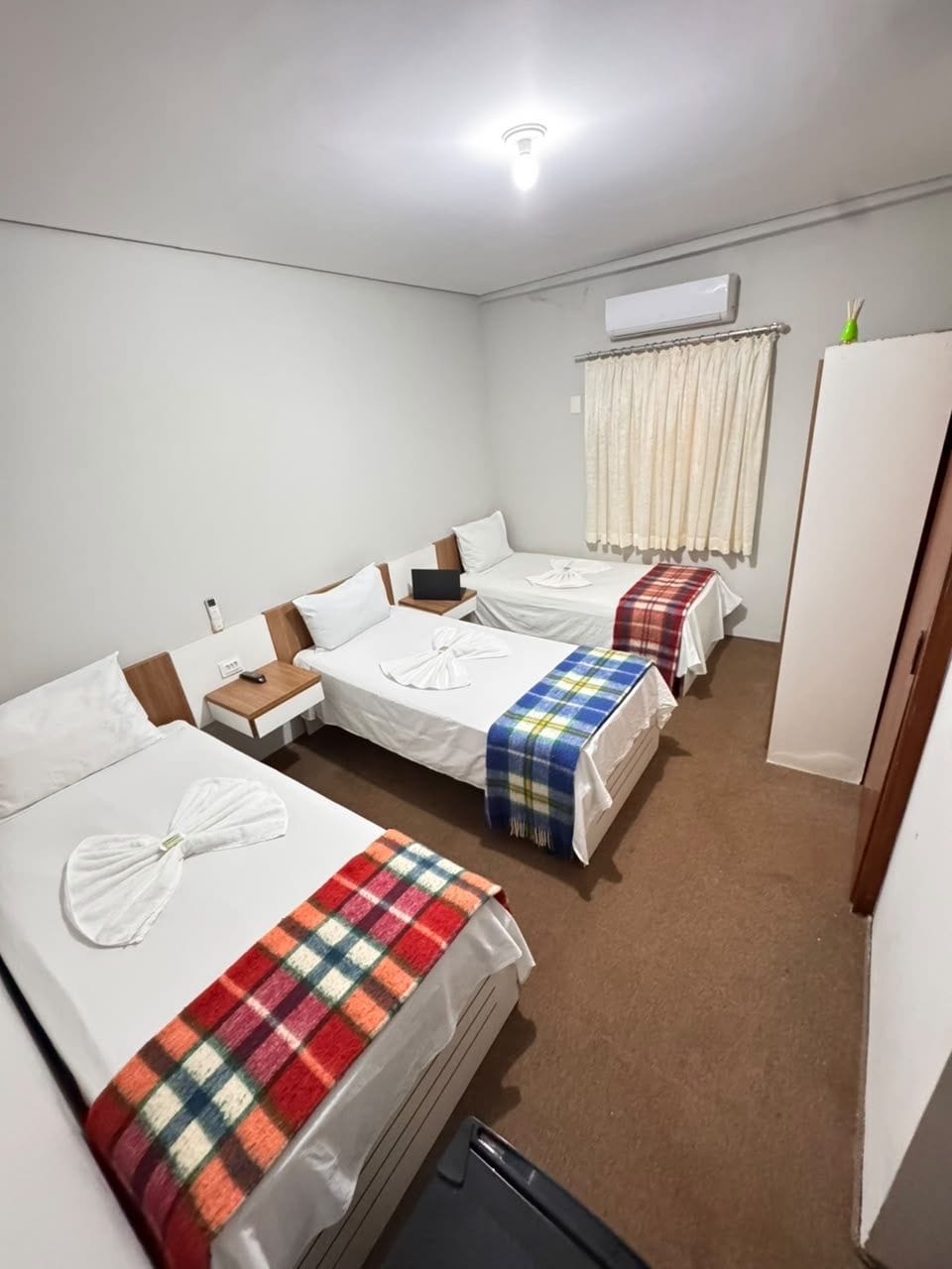 Quarto de hotel com duas camas, mesa de cabeceira, guarda-roupa e ar condicionado.