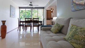 Uma sala de estar moderna com um sofá cinzento, uma mesa de jantar com cadeiras, um ventilador de teto e uma grande janela com vista para o verde.