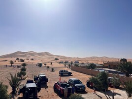Paisagem desértica com dunas de areia, veículos estacionados e vegetação rala.