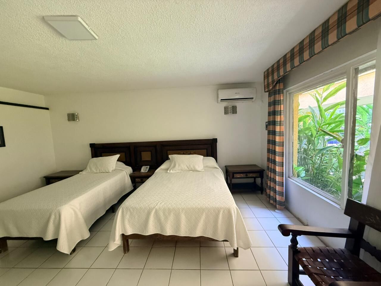 Quarto com duas camas de solteiro, um banco, uma janela com cortinas e um ar condicionado.