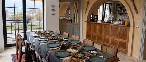 Uma sala de jantar com uma mesa comprida posta para refeição, uma cozinha com armários de madeira e vista através de grandes janelas.