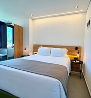 Quarto de hotel moderno com uma cama grande, cabeceira de madeira, mesa de cabeceira com abajur e vista da cidade através de uma janela ampla.