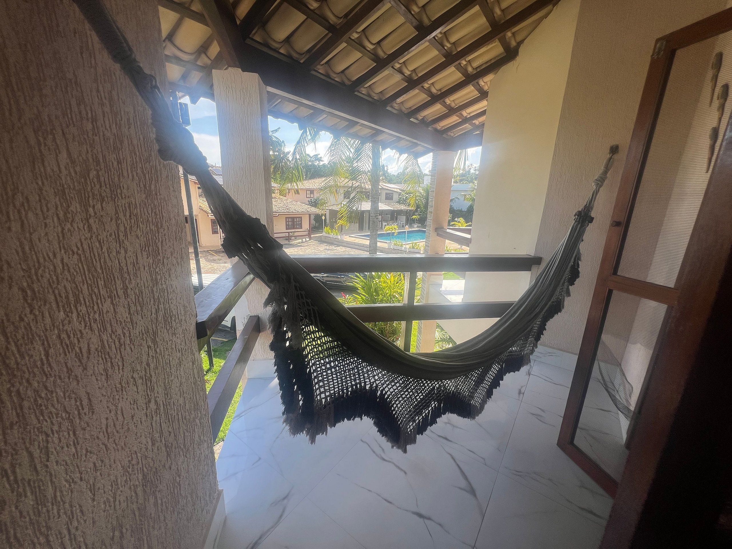 Uma rede estendida entre dois pilares em área externa coberta, com vista para uma piscina e edifícios ao fundo.