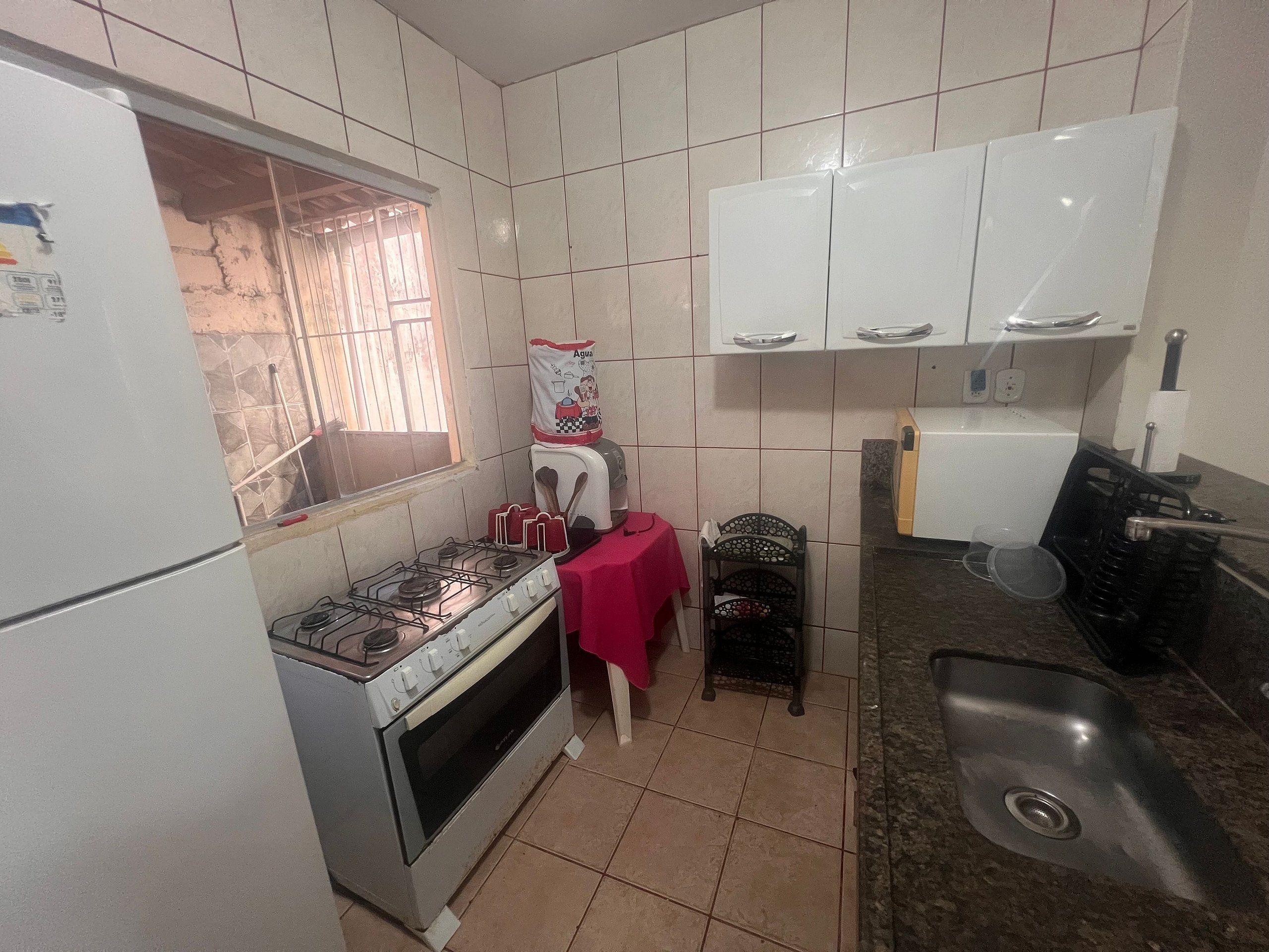Cozinha compacta com armários brancos, fogão a gás, pia e janela com grades.