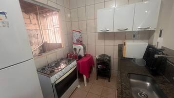Cozinha compacta com armários brancos, fogão a gás, pia e janela com grades.