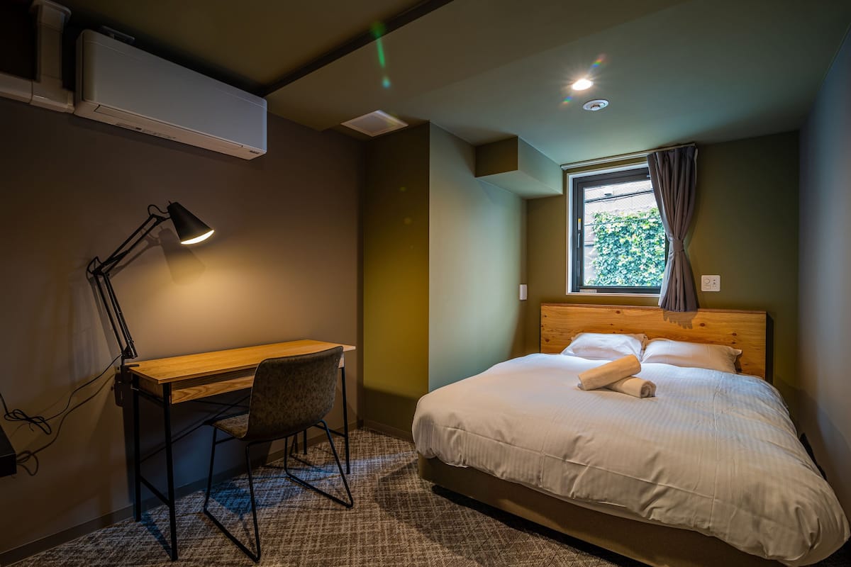 Quarto com cama, escrivaninha, cadeira, janela com cortinas e ar condicionado.