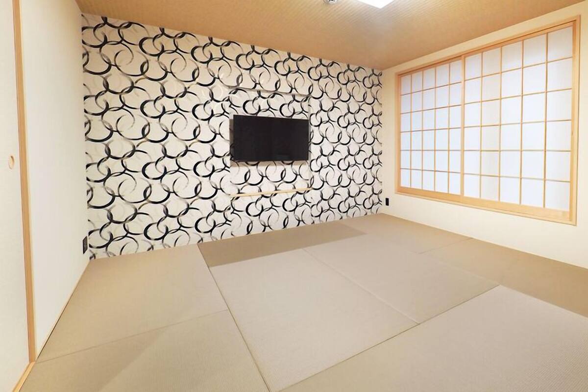 Quarto com piso de tatame, uma TV preta fixada numa parede estampada e uma porta de papel shoji.