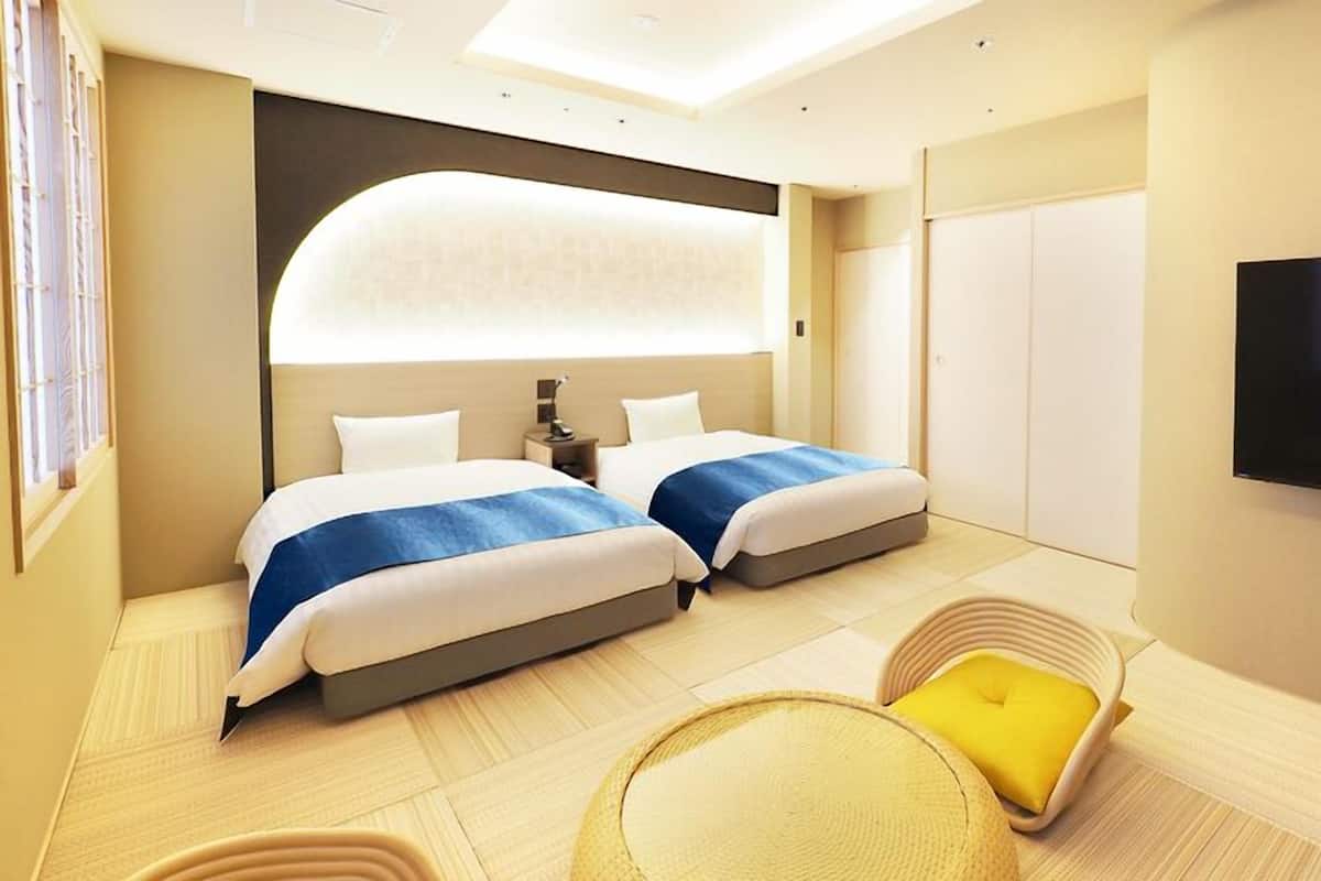 Quarto de hotel com duas camas, televisão de tela plana, pequeno espaço de estar e uma janela grande.