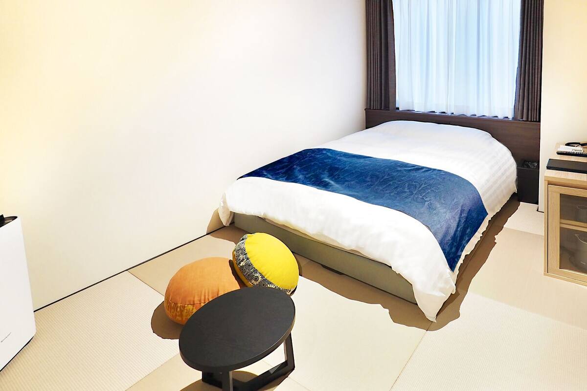 Quarto de hotel com cama, mesinha, cadeira, minibar e janela com cortinas.
