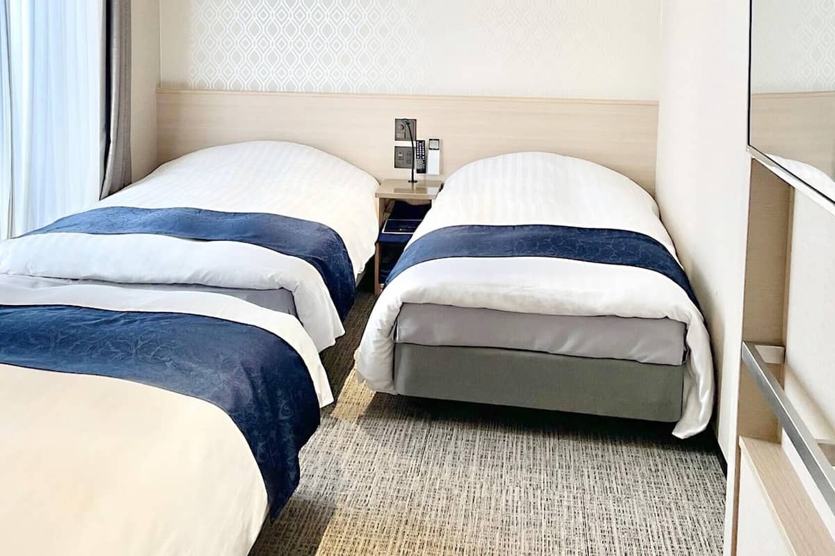 Quarto de hotel com duas camas, uma mesa de cabeceira com telefone e abajur, e uma parede com padrão.