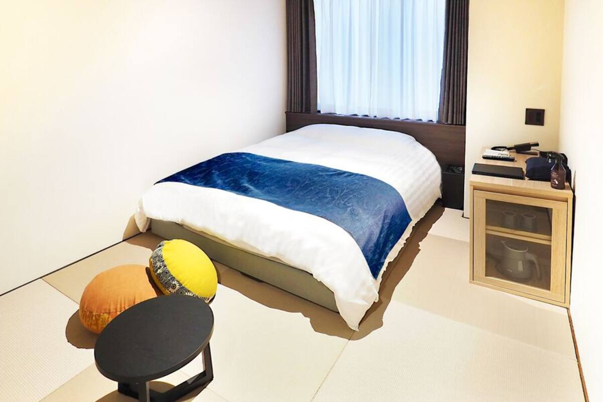 Quarto de hotel com cama, uma mesinha, uma cadeira, mesa de cabeceira com abajur e janela com cortinas.