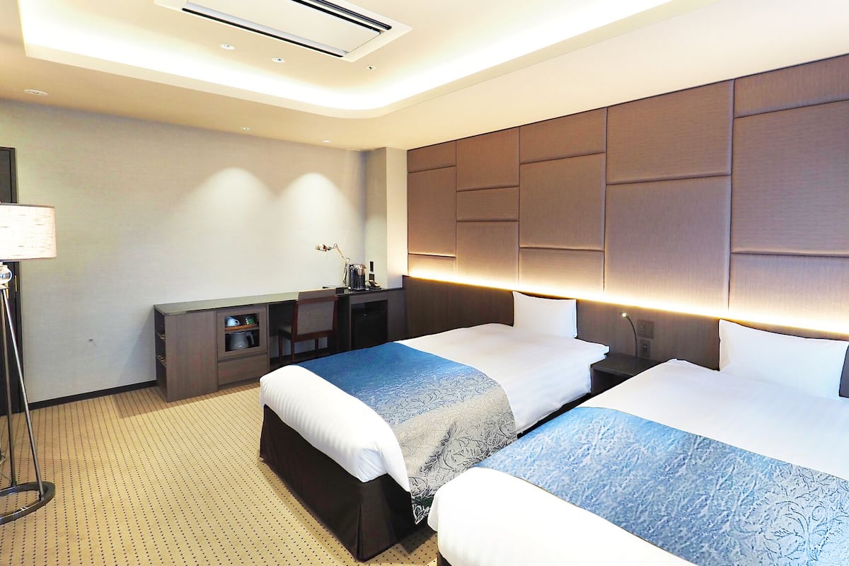 Quarto de hotel com duas camas, uma escrivaninha, uma luminária e um respiradouro no teto.