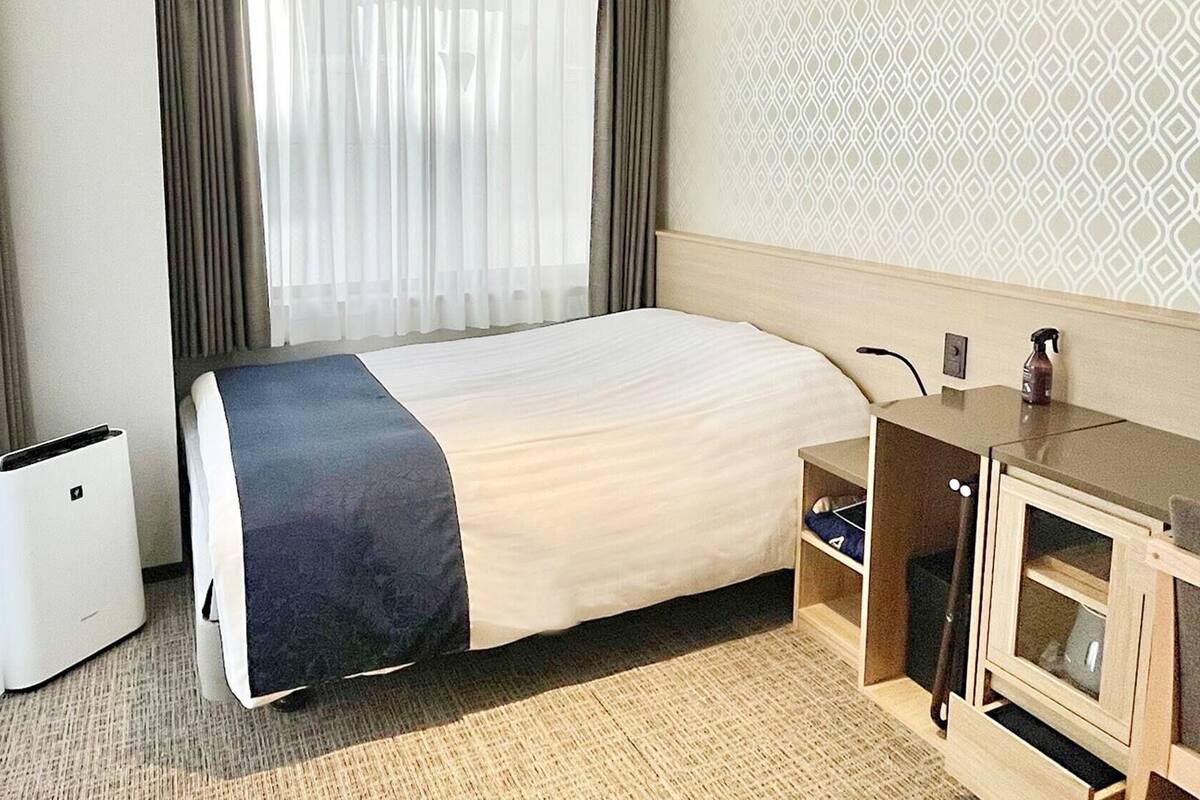 Quarto de hotel com uma cama, escrivaninha, cadeira, armário pequeno e lixeira.