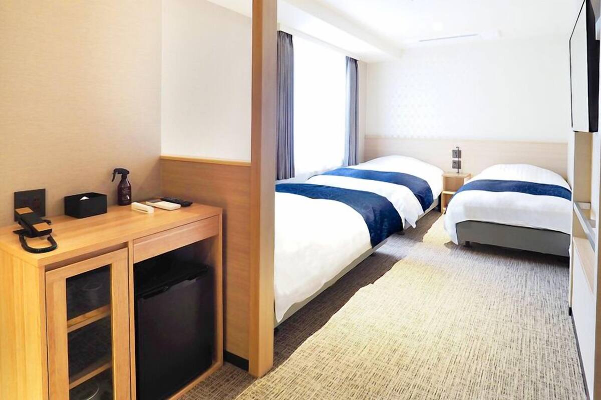 Quarto de hotel com secretária de madeira, duas camas, televisão e janela com cortinas.