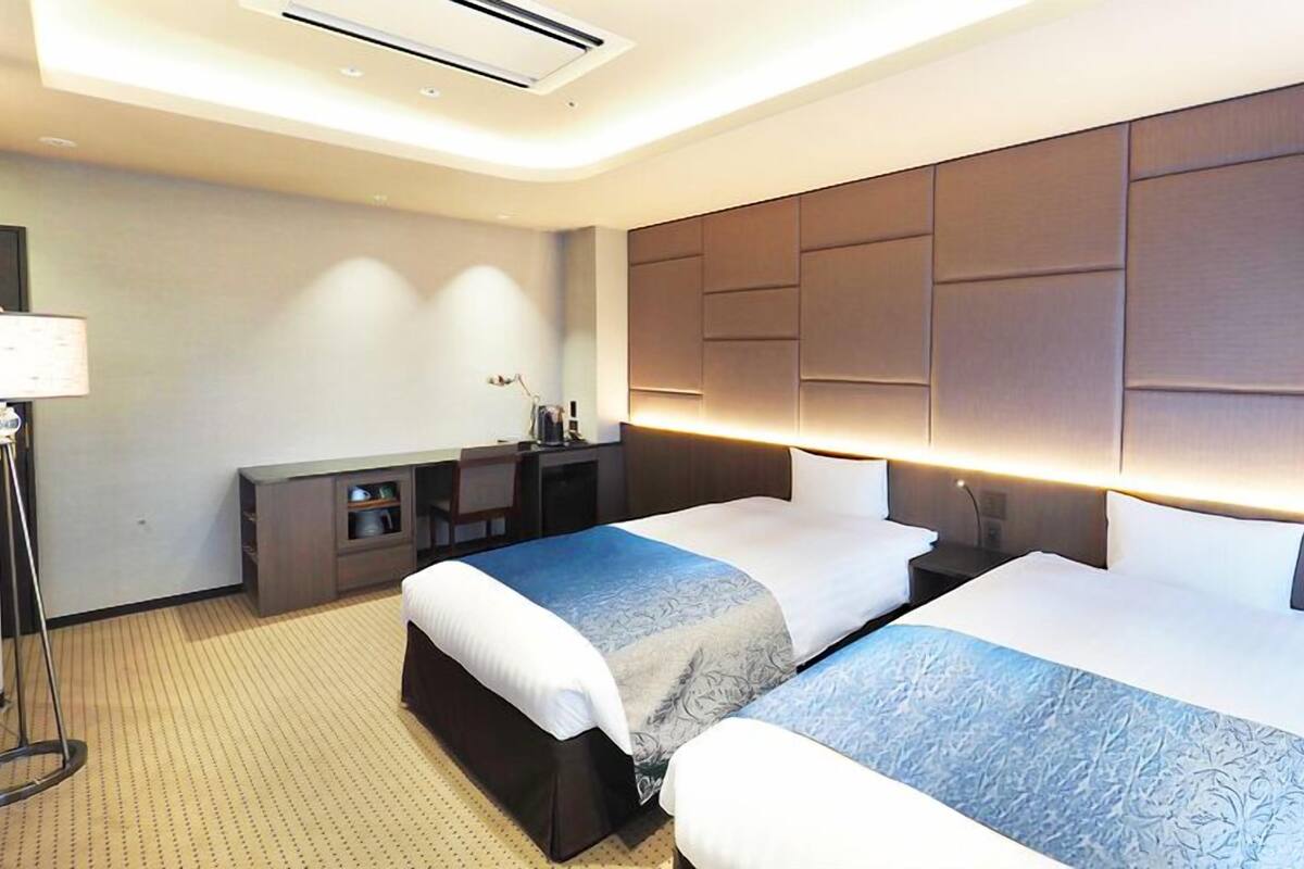 Quarto de hotel com duas camas, uma escrivaninha, uma luminária e um respiradouro no teto.
