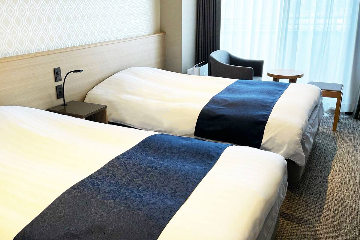 Quarto de hotel com duas camas, mesa de cabeceira, cadeira, mesa pequena e janela com cortinas.