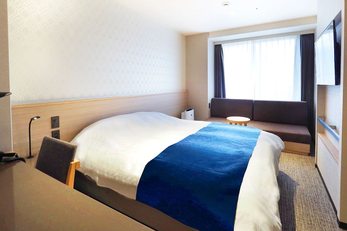 Quarto de hotel com uma cama, uma mesinha, um sofá, uma luminária e uma janela com cortinas.