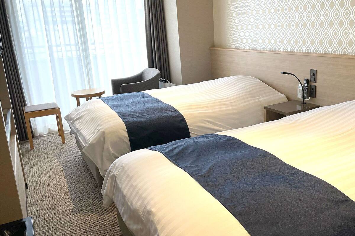 Quarto de hotel com duas camas, uma escrivaninha, uma cadeira e um papel de parede estampado.