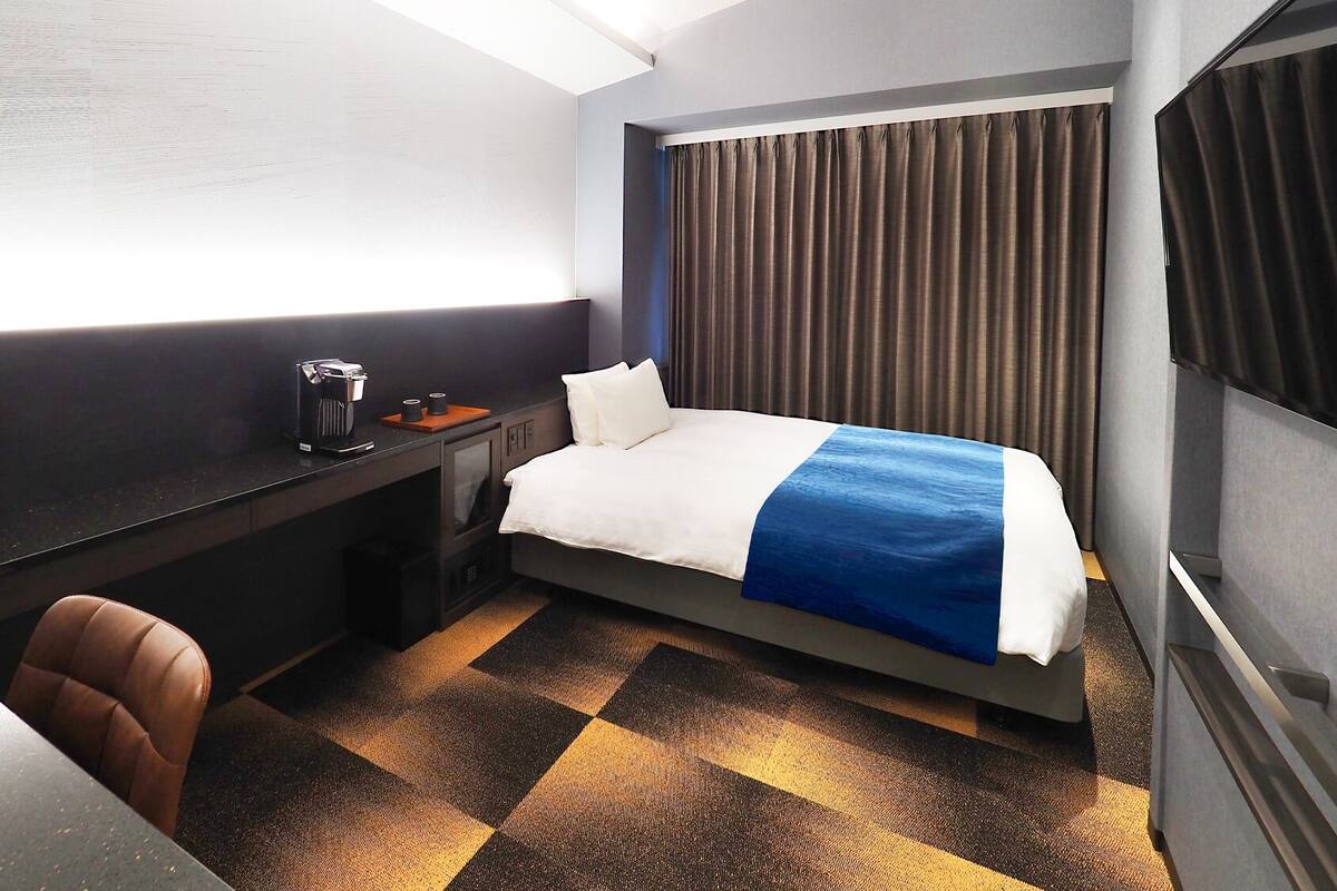 Quarto de hotel com cama, escrivaninha, cadeira, cafeteira e janela com cortinas.