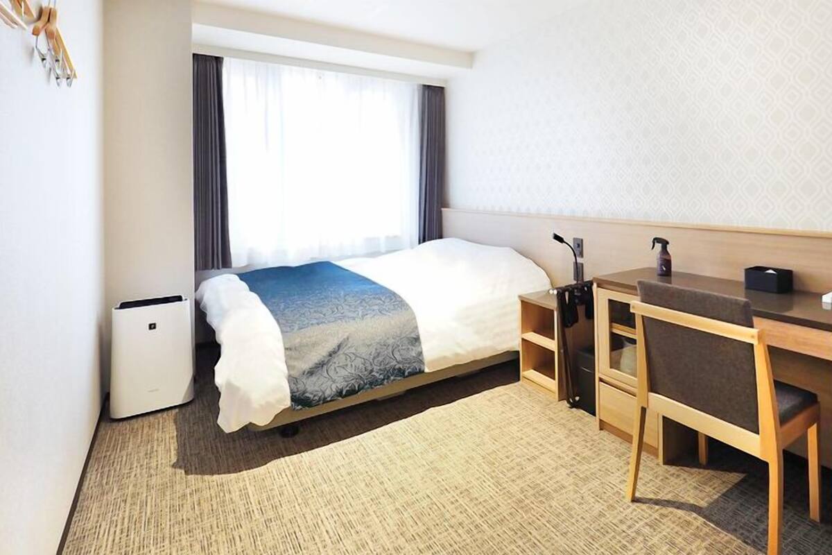 Quarto de hotel com cama, escrivaninha, cadeira e um pequeno aparelho de ar condicionado.