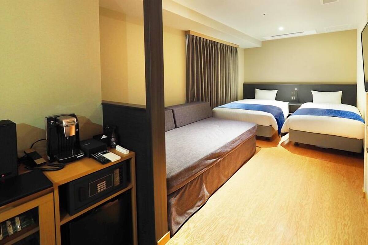 Quarto de hotel com escrivaninha, duas camas, um sofá, televisão e cafeteira.
