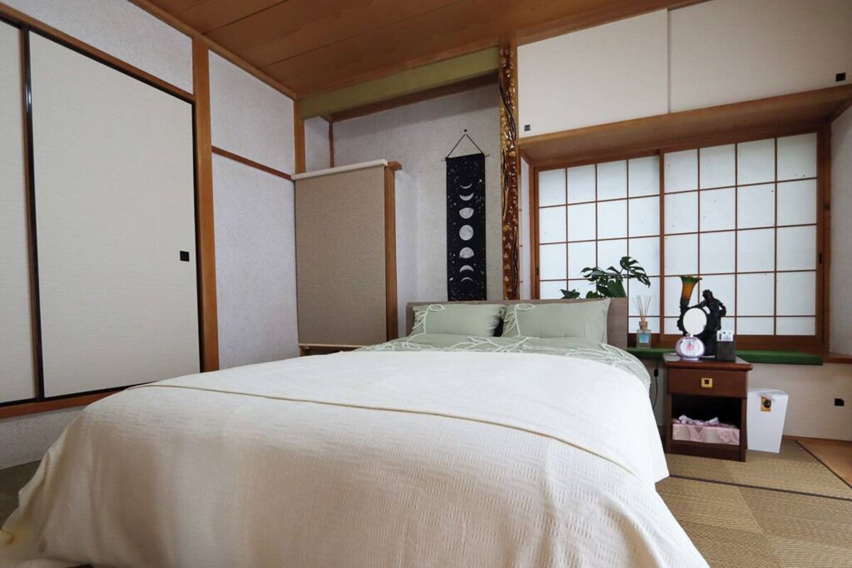 Um quarto japonês tradicional com tatame, uma cama baixa com colcha branca, um biombos shoji e um pergaminho pendurado.