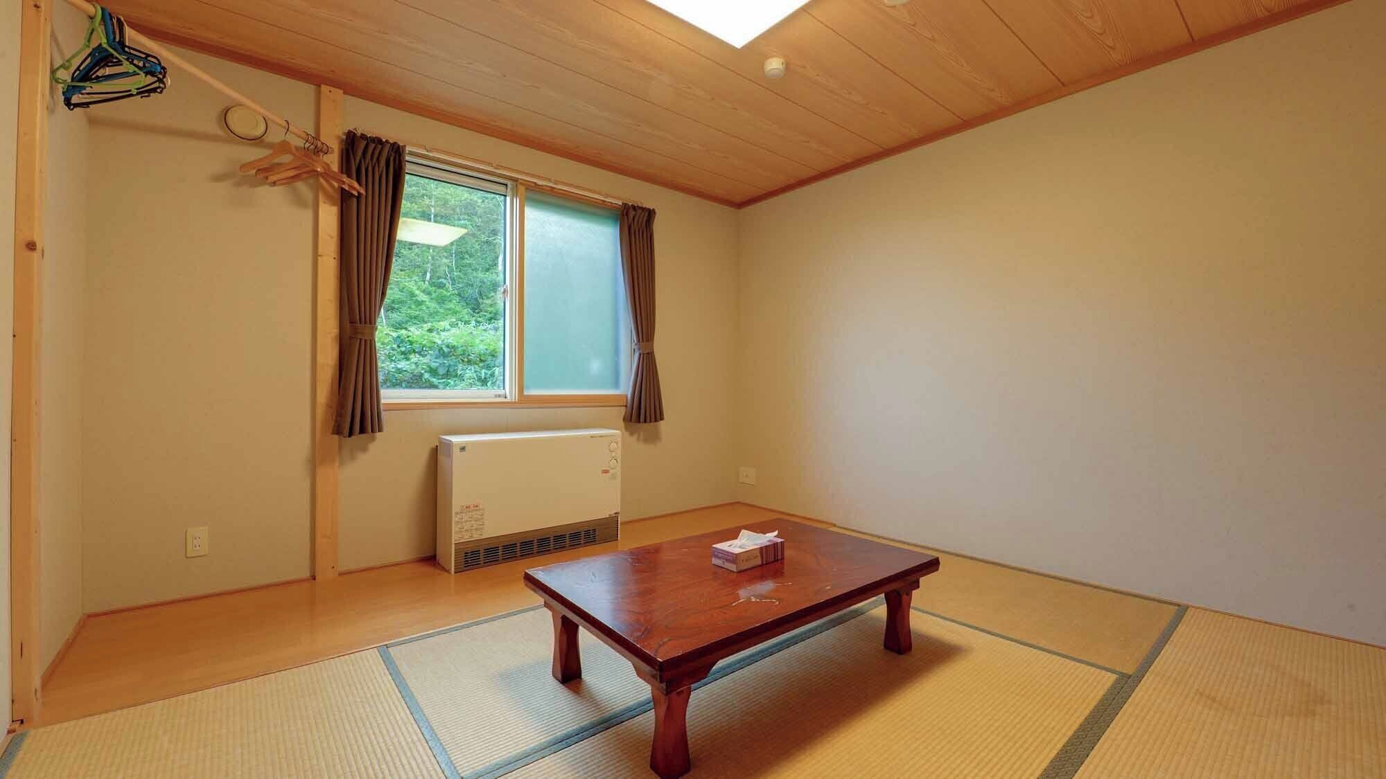 Quarto com teto de madeira, janela com cortinas, radiador, piso de esteira de tatame e mesa de madeira com caixa de lenços.