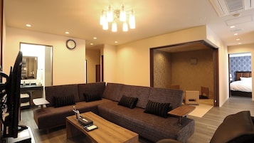 Quarto de hotel moderno com um amplo sofá de canto, uma mesa de centro de madeira, uma televisão e um banheiro visível por uma porta aberta.