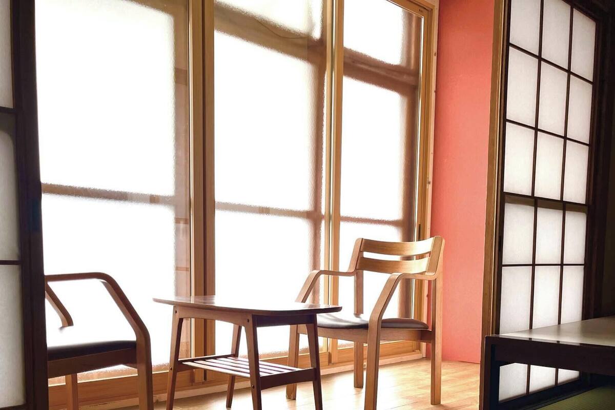 Quarto com mesa de madeira, duas cadeiras, janela com vista e pequena mesa com caixa de lenços.