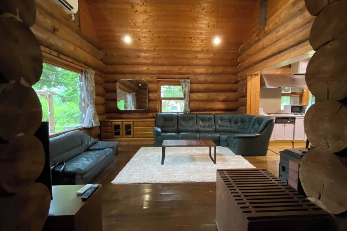 Interior de uma cabana de tronco com sofá, mesa de centro, área de cozinha e uma janela grande.