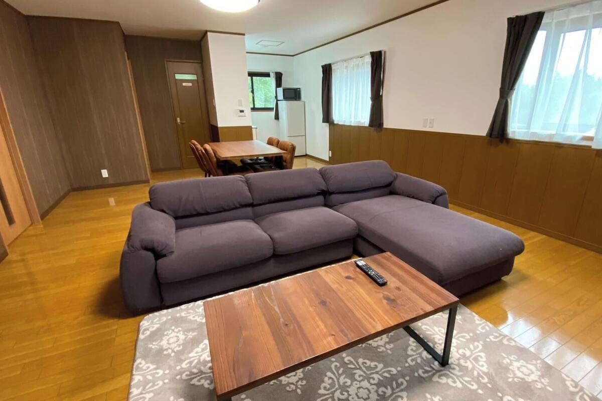 Uma sala de estar com um sofá seccional, uma mesa de centro de madeira e uma área de jantar ao fundo.