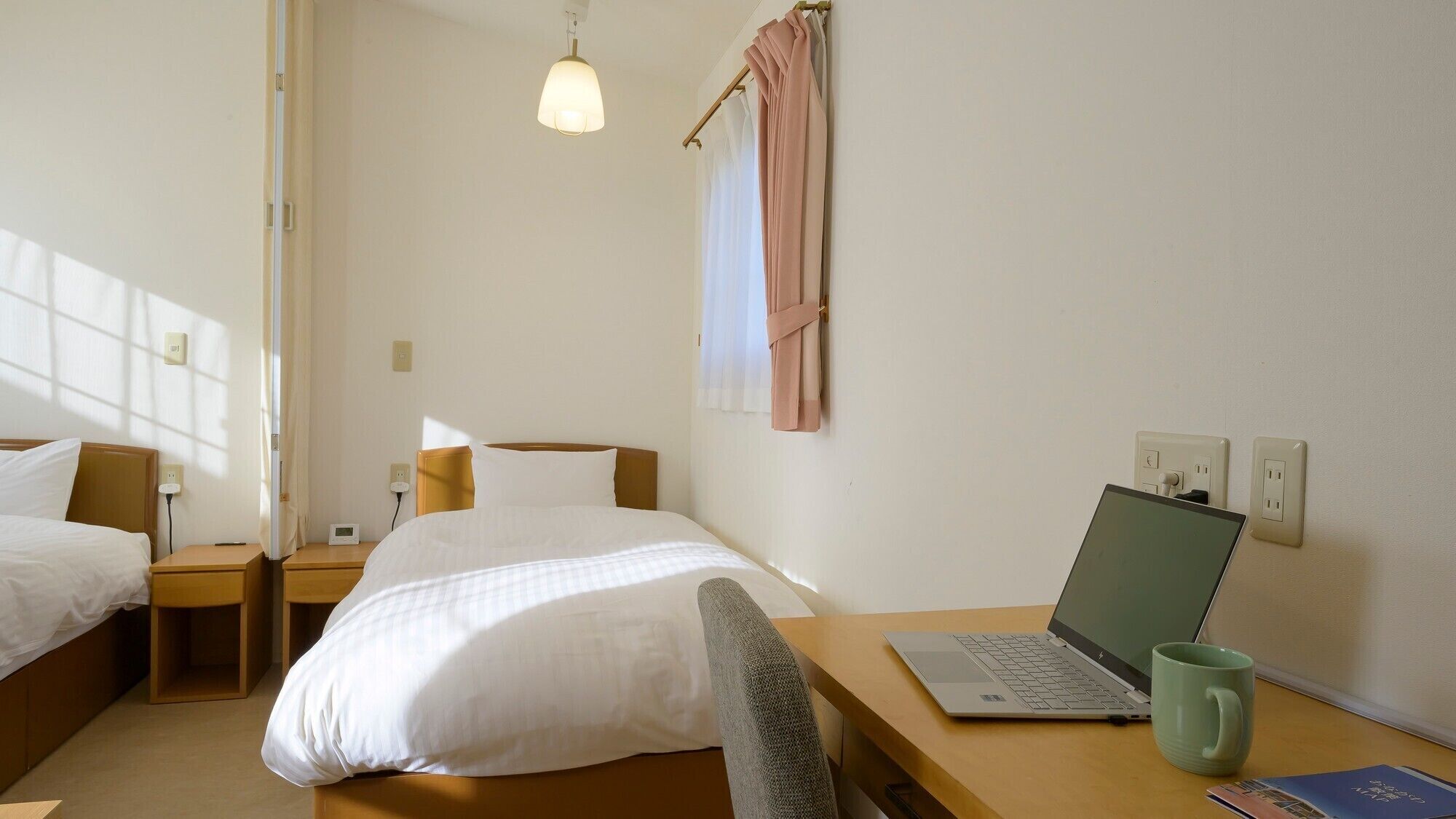 Quarto de hotel com duas camas, uma escrivaninha com um laptop, uma caneca e uma janela com cortinas.