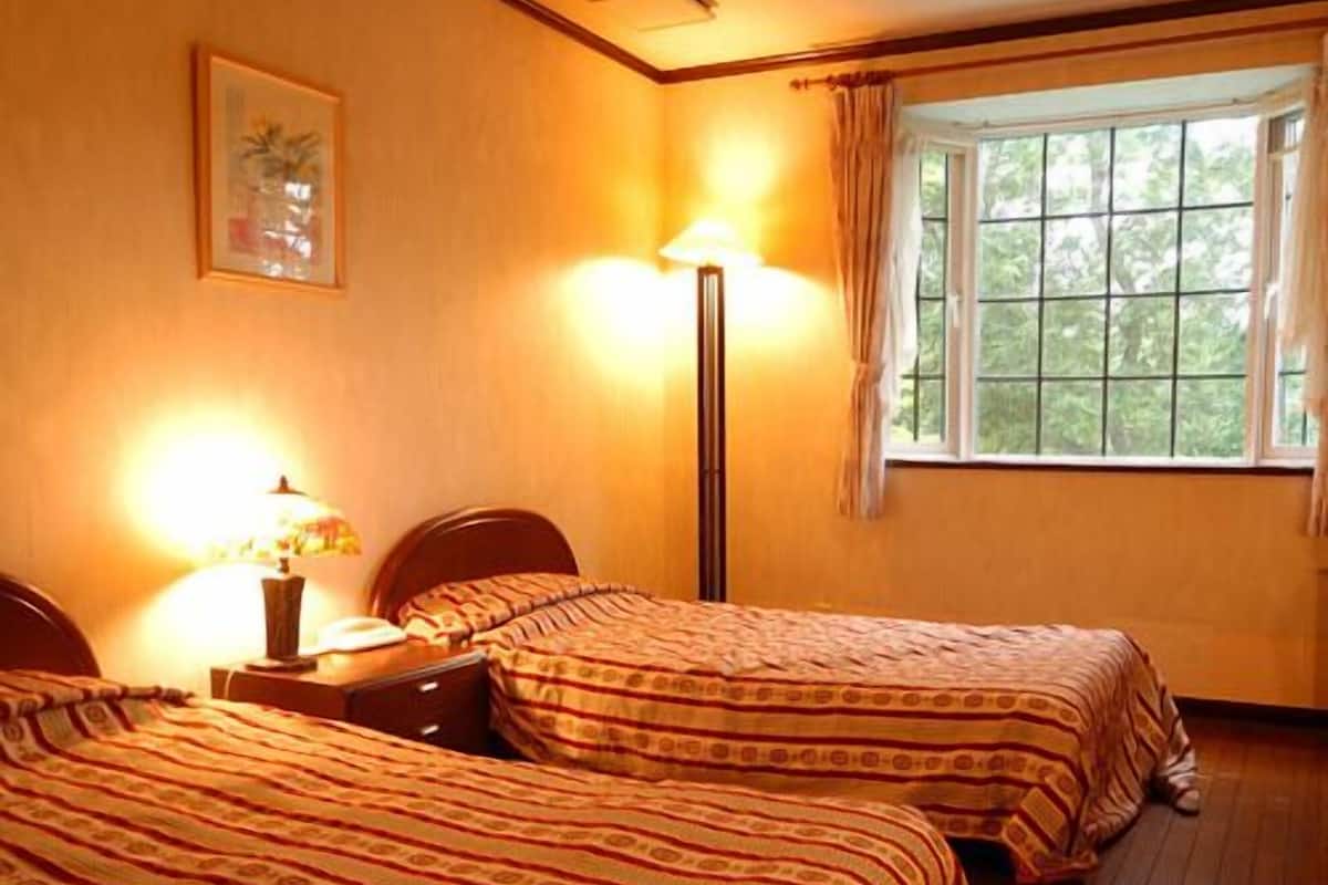 Quarto com duas camas, uma mesinha de cabeceira, uma luminária, uma janela com cortinas e um quadro na parede.
