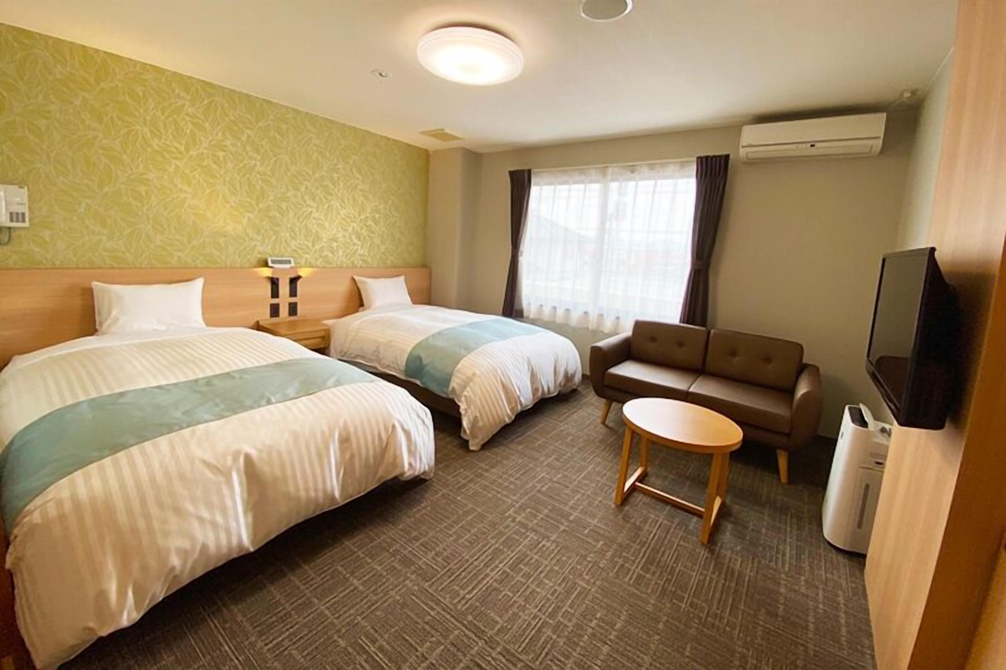 Quarto de hotel com duas camas, um sofá, uma mesa pequena, uma televisão e um ar condicionado.