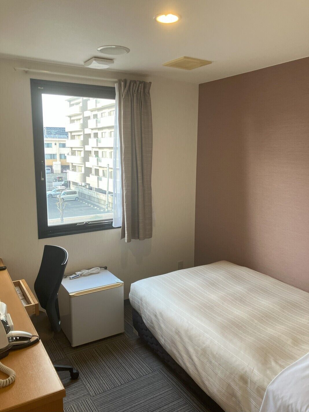 Quarto de hotel com cama, escrivaninha, cadeira e janela com vista para edifícios.
