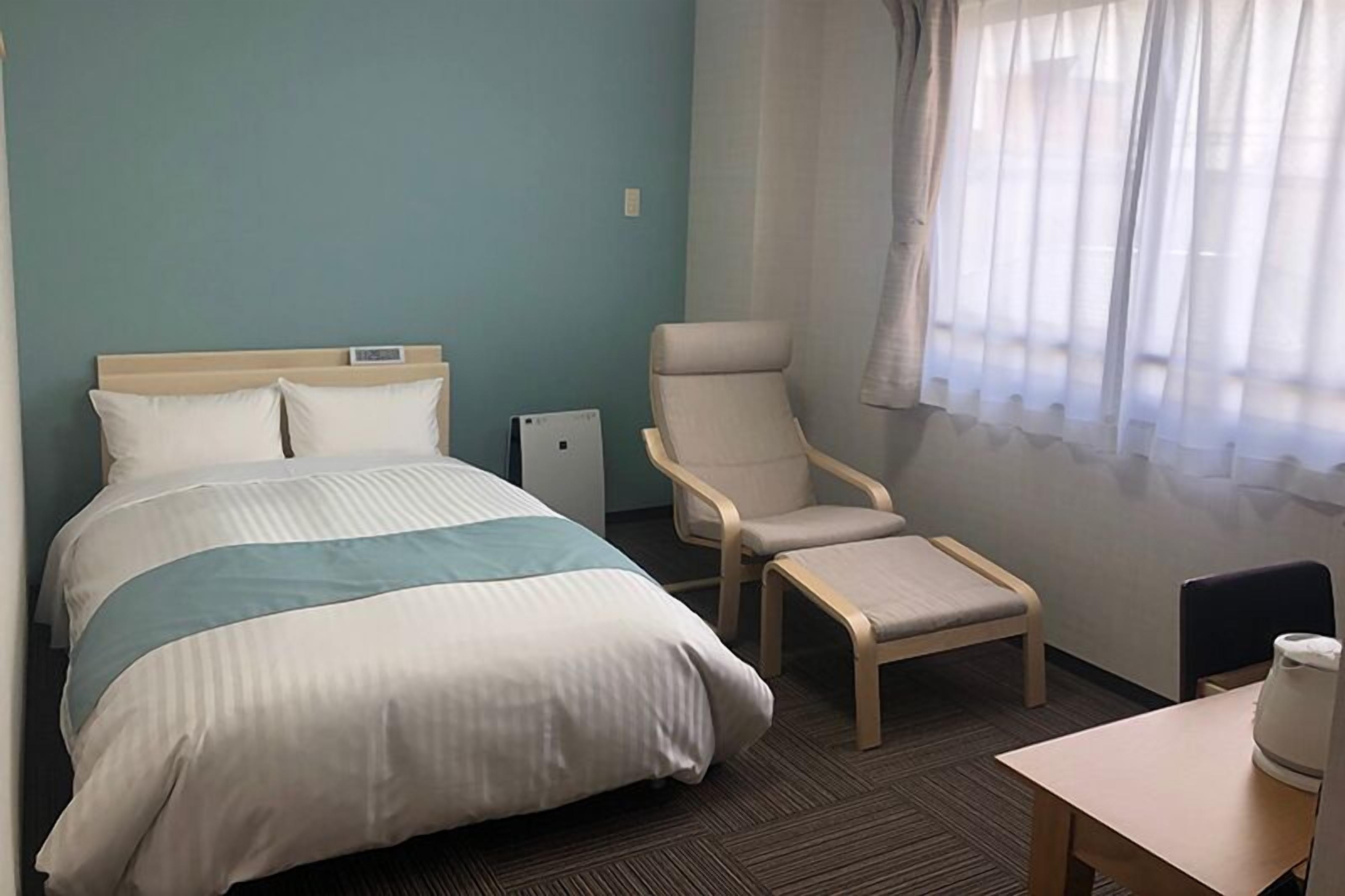 Quarto de hotel com cama, uma cadeira, uma mesinha e janela com cortinas.