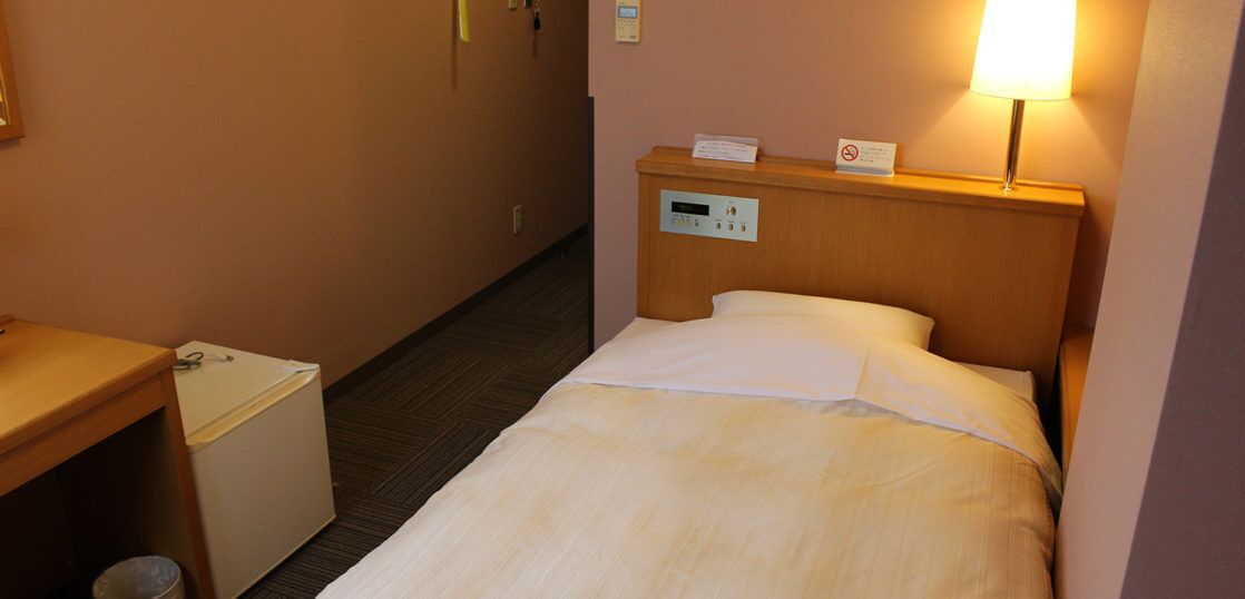 Quarto de hotel com uma cama, uma escrivaninha, um cesto de lixo e um painel de controle fixado na parede.