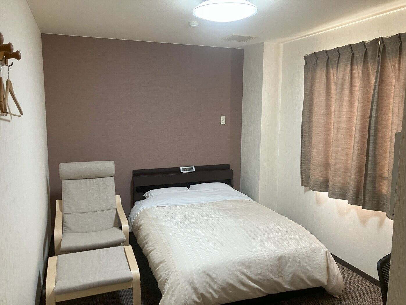 Uma cama de solteiro com cabeceira, uma cadeira, um relógio de parede e uma janela com cortinas.