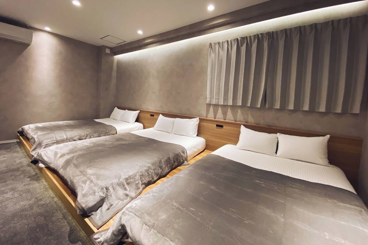 Quarto de hotel com três camas dispostas em linha, uma janela grande com cortinas e design moderno.