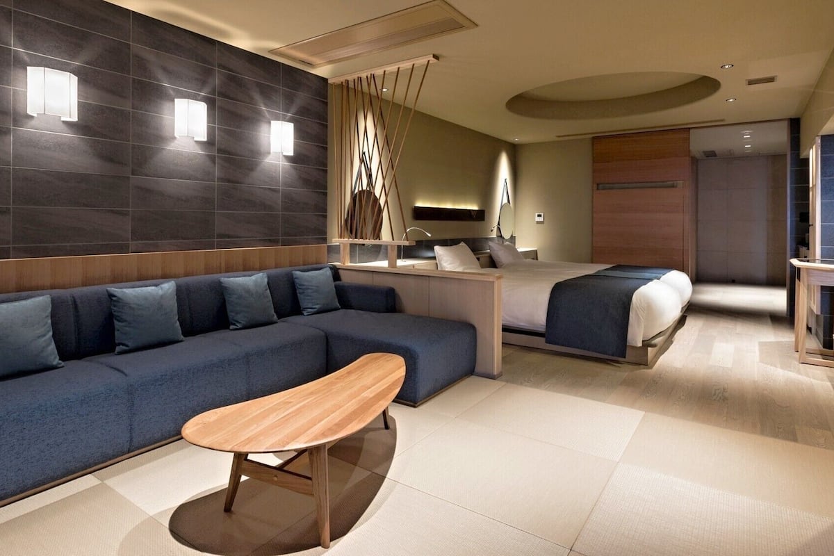 Quarto de hotel moderno com um amplo sofá modular, uma mesa de centro de madeira, uma cama, uma escrivaninha e uma janela grande.