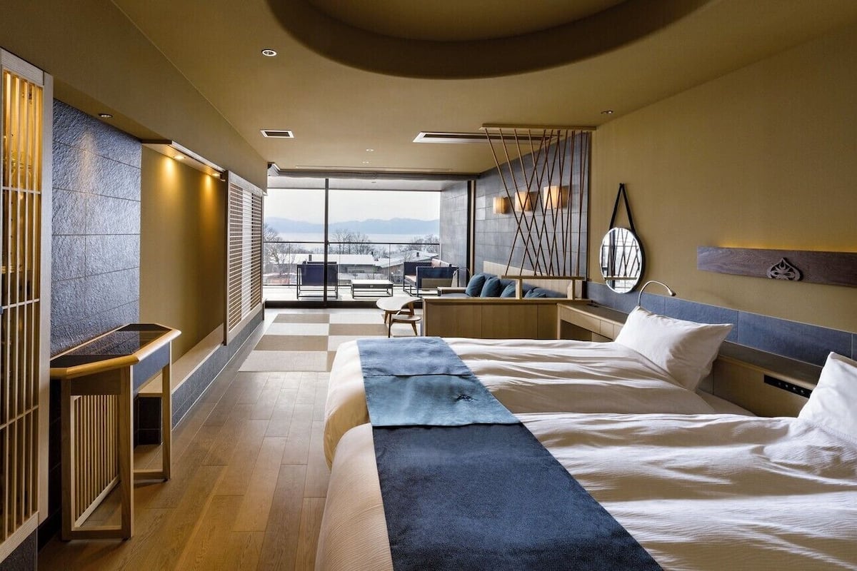 Quarto de hotel com uma cama grande, uma escrivaninha, uma cadeira, vista para o exterior e um detalhe decorativo na parede.