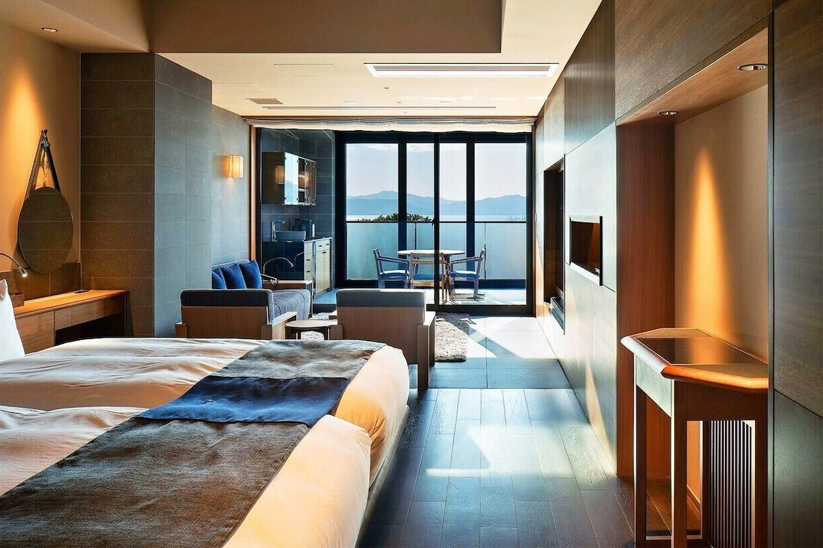 Quarto de hotel moderno com uma cama grande, uma mesa, uma cadeira, uma mesinha e vista para as montanhas através de uma janela ampla.