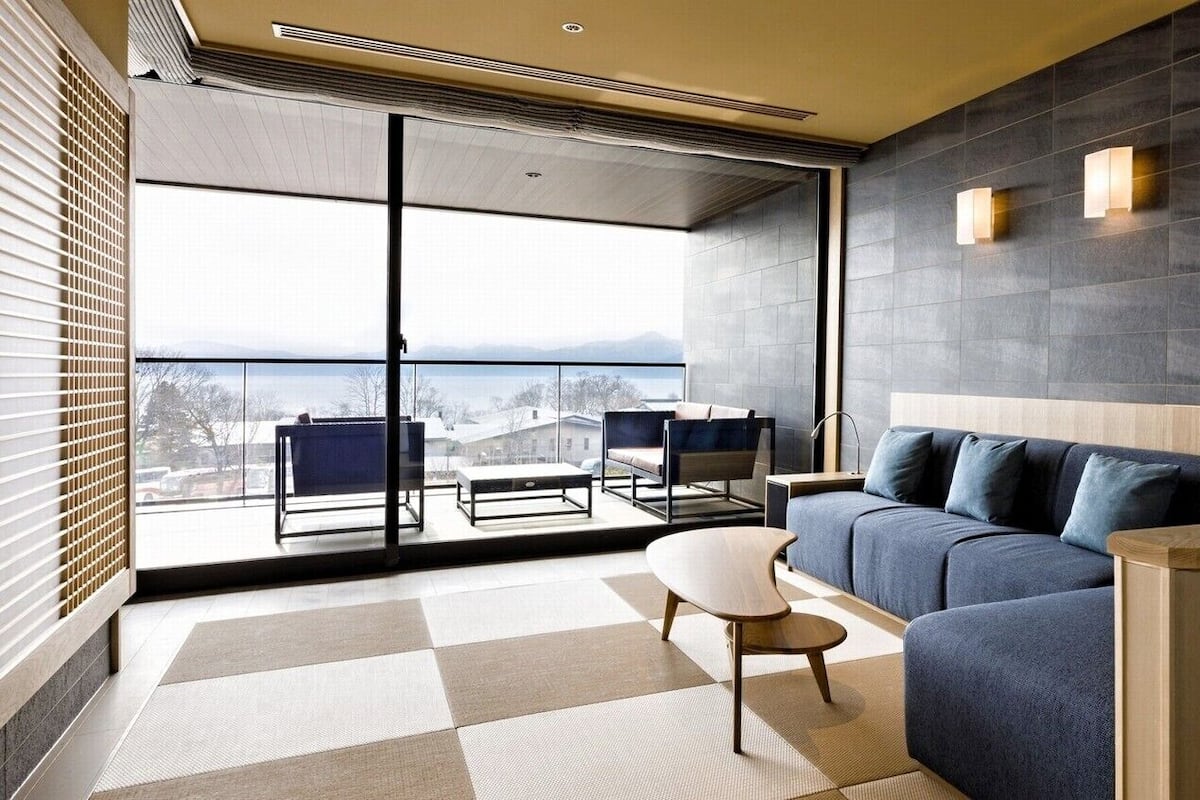Quarto de hotel moderno com uma varanda grande, um sofá, uma mesa de centro e vista para a paisagem urbana.