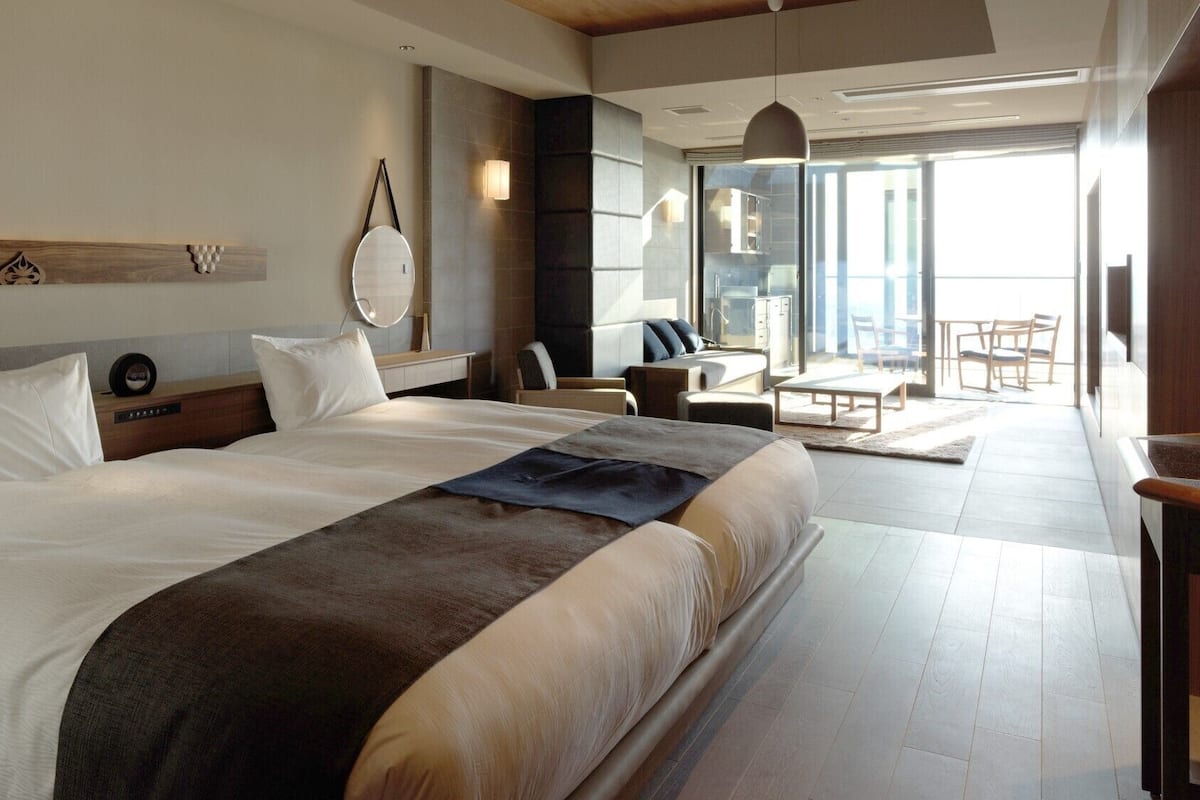 Quarto de hotel moderno com uma cama grande, área de estar com sofá e cadeiras, e varanda com vista.
