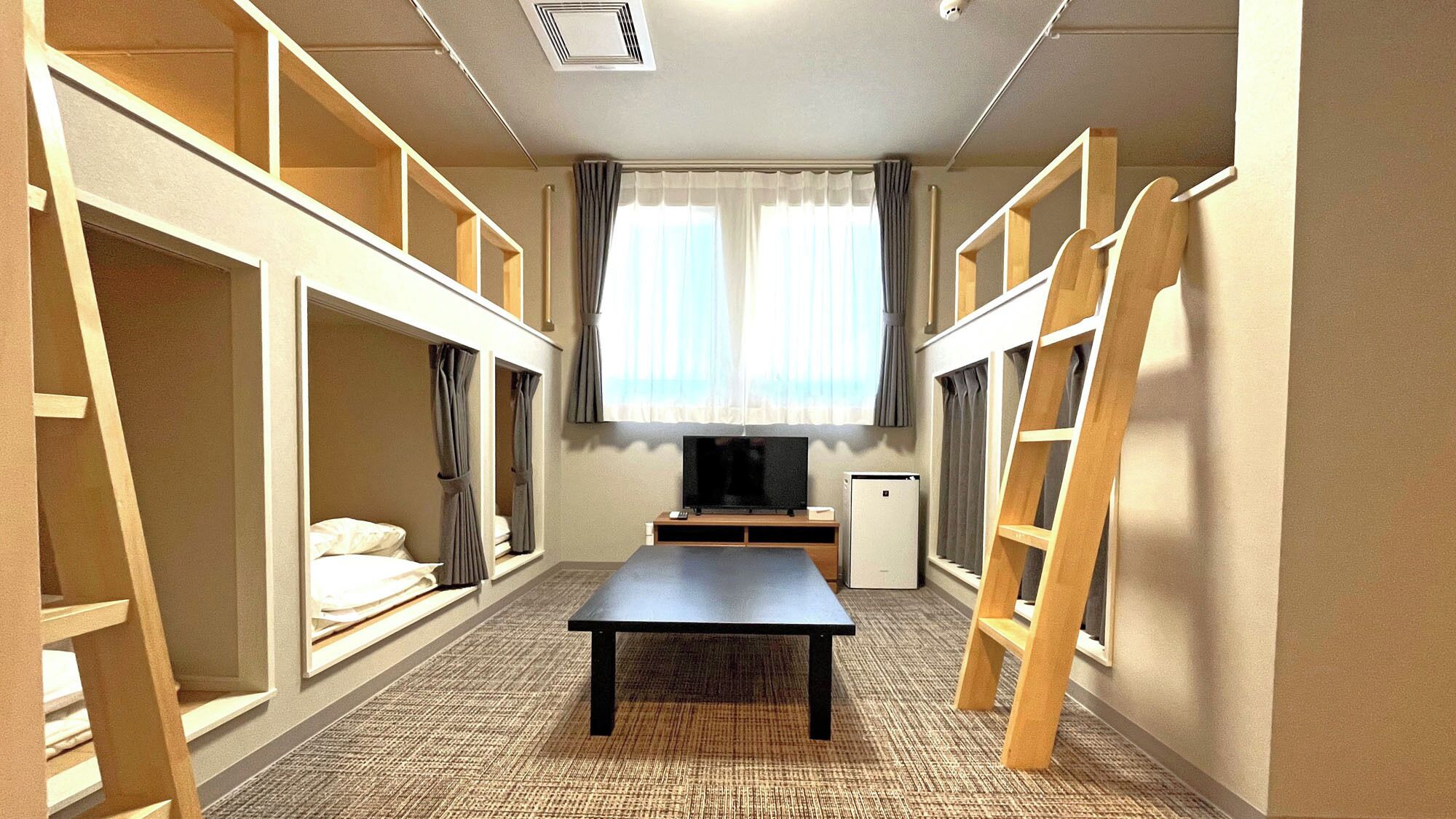 Quarto com beliches, televisão, uma mesinha e janela com cortinas.